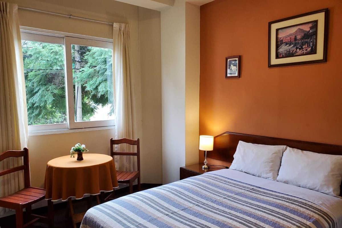 Quarto de hotel com uma cama grande, uma mesa redonda com cadeiras, uma janela com cortinas e um quadro na parede.