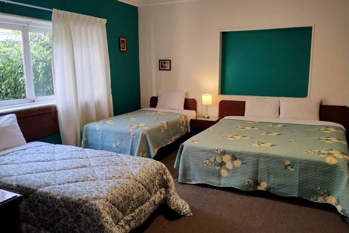 Quarto de hotel com duas camas, janela com cortinas, parede de destaque verde e mesa de cabeceira com abajur.