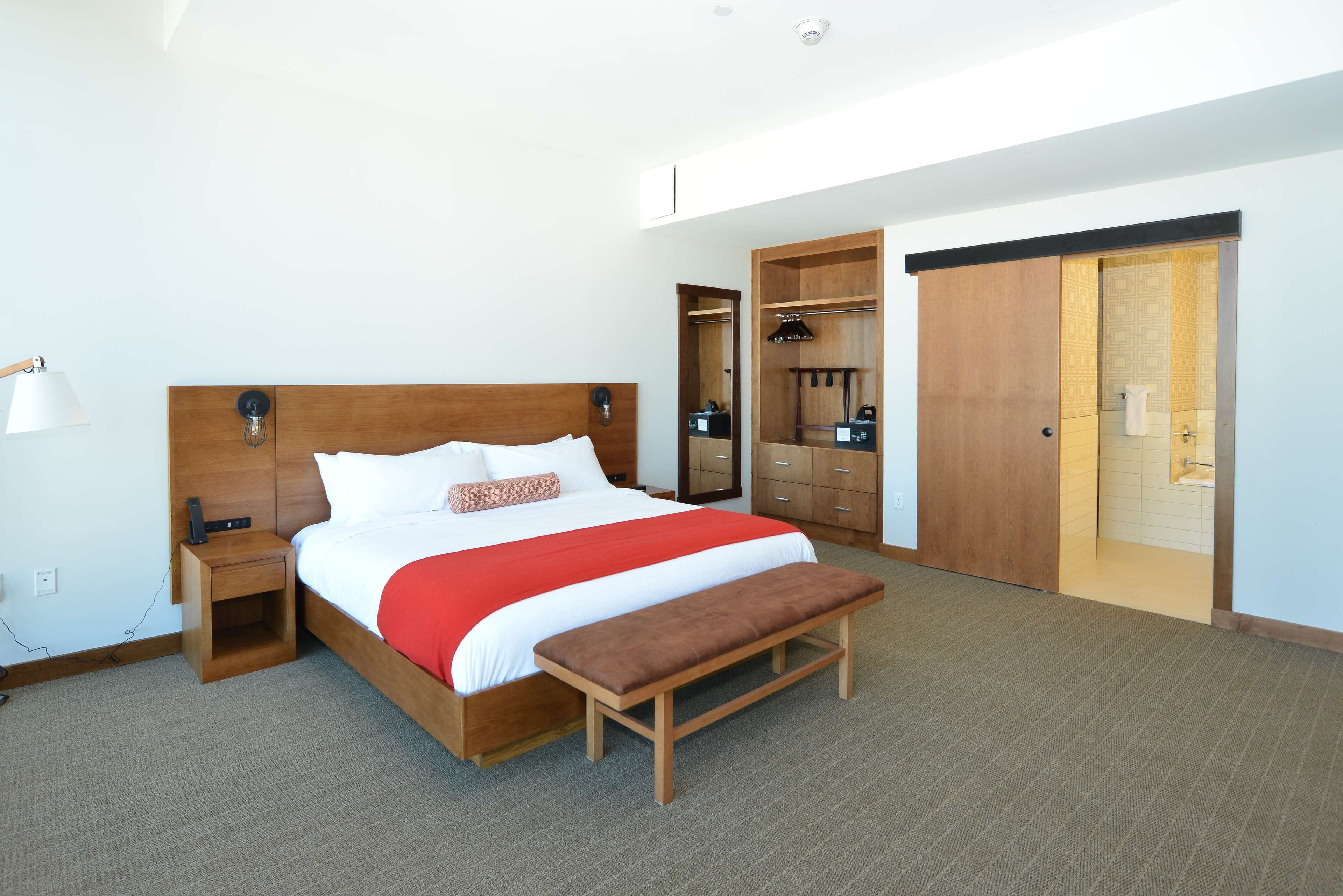 Quarto de hotel moderno com uma cama grande, cabeceira de madeira, um banco, uma mesinha de cabeceira, um guarda-roupa e um closet espaçoso.