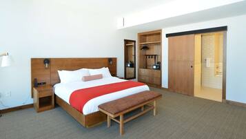 Quarto de hotel moderno com uma cama grande, cabeceira de madeira, um banco, uma mesinha de cabeceira, um guarda-roupa e um closet espaçoso.