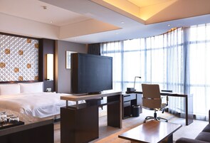 Quarto de hotel moderno com cama, escrivaninha, cadeira, televisão e grandes janelas.