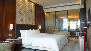 Quarto de hotel moderno com cama grande, divisória de vidro, banheiro com espelho e porta-toalhas vermelho.