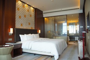 Quarto de hotel moderno com cama grande, divisória de vidro, banheiro com espelho e porta-toalhas vermelho.