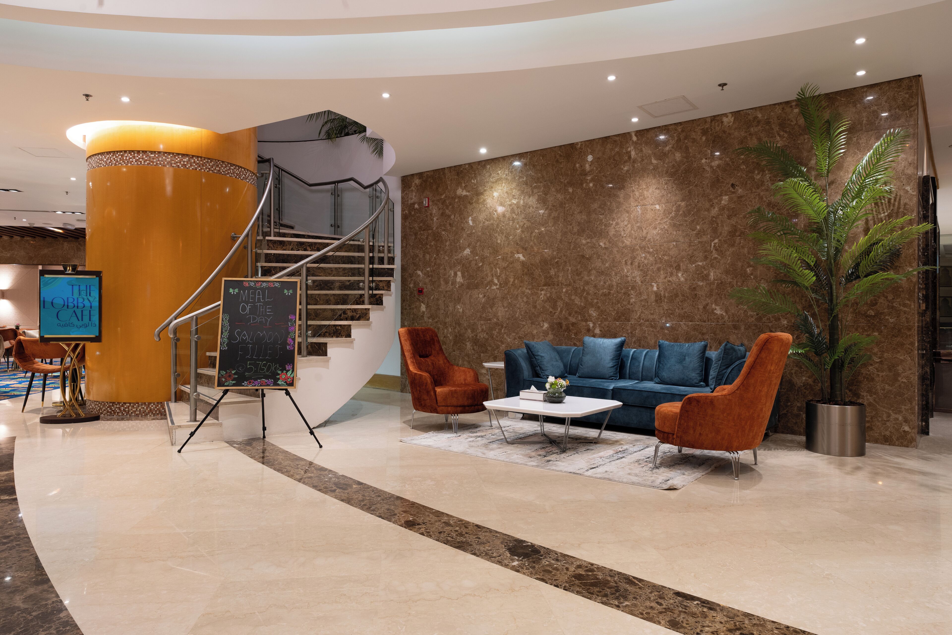 Um lobby moderno com piso de mármore, escadaria, sofá azul, poltrona laranja e uma grande planta em vaso.