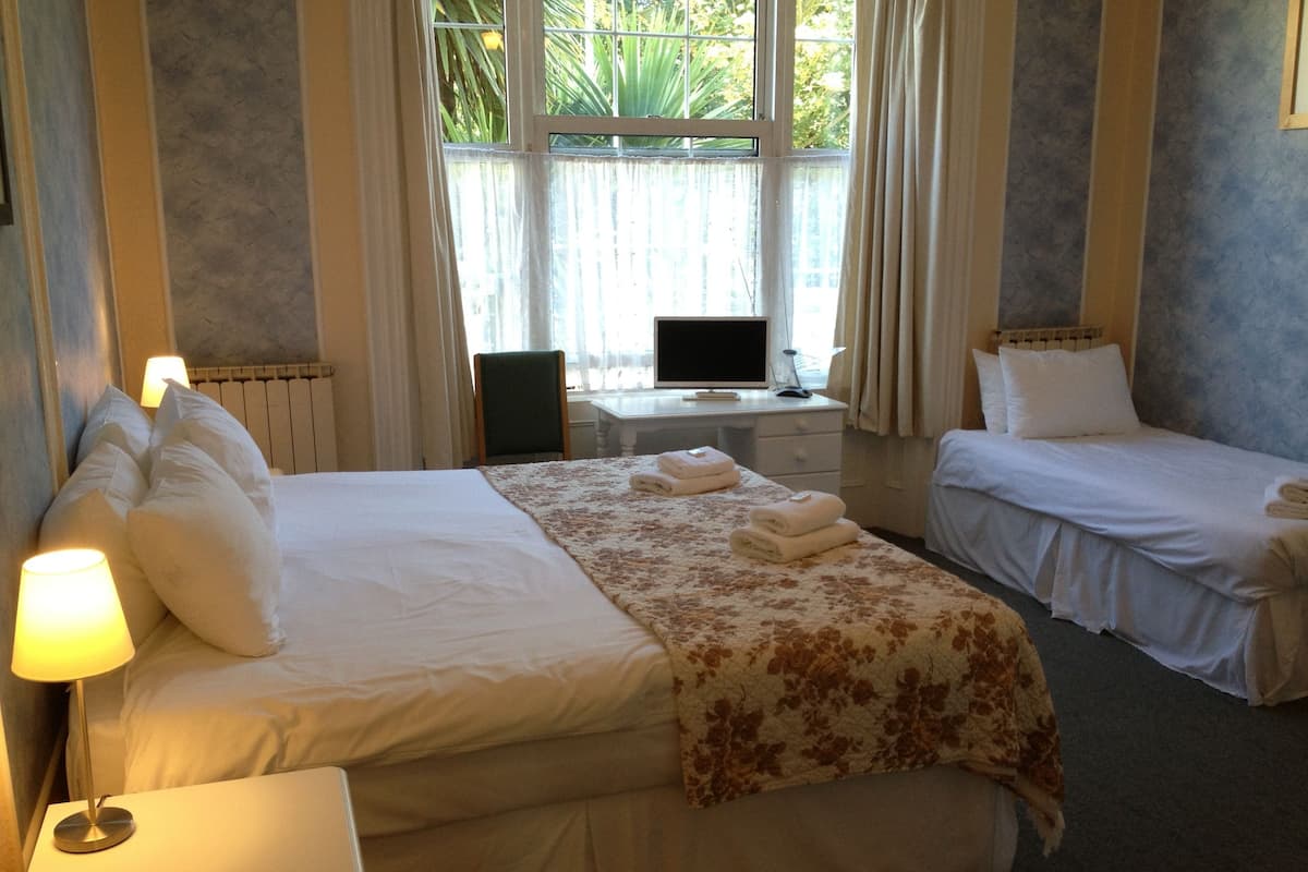 Quarto de hotel com uma cama grande, uma cama menor, uma escrivaninha com computador e uma janela com cortinas.