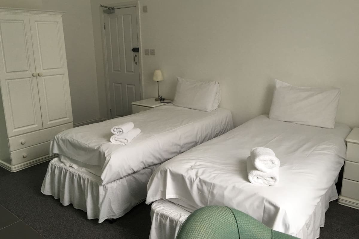 Quarto de hotel com duas camas de solteiro, um guarda-roupa, uma mesa de cabeceira com abajur e uma cadeira verde.