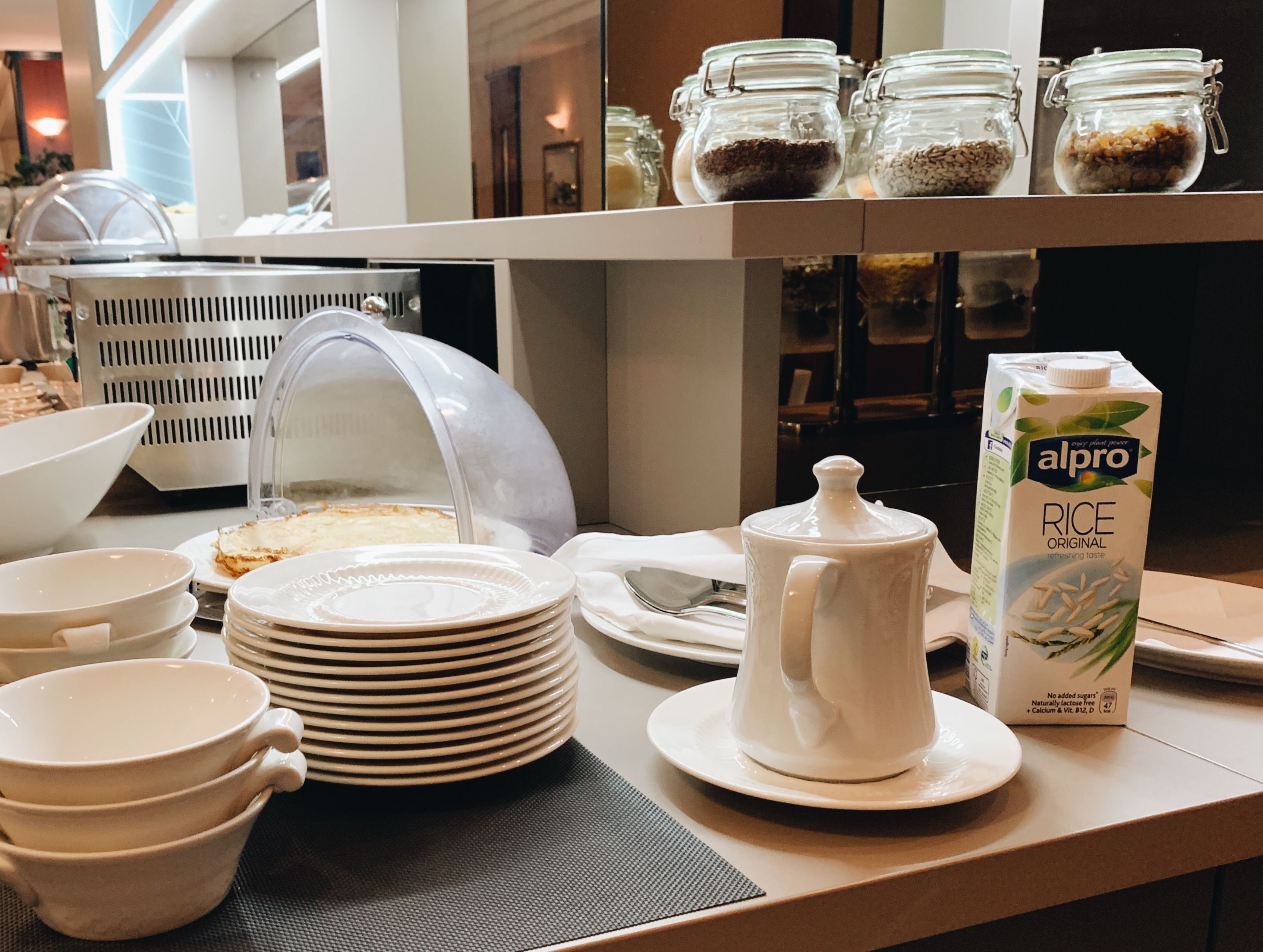 Buffet de café da manhã com pratos e tigelas empilhados, aquecedor de alimentos coberto e um recipiente com leite de arroz.