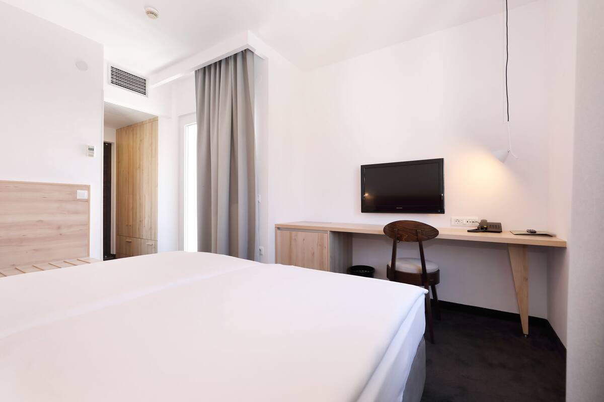 Quarto de hotel com uma cama grande, uma mesa de escritório com um computador, uma televisão e uma cadeira.