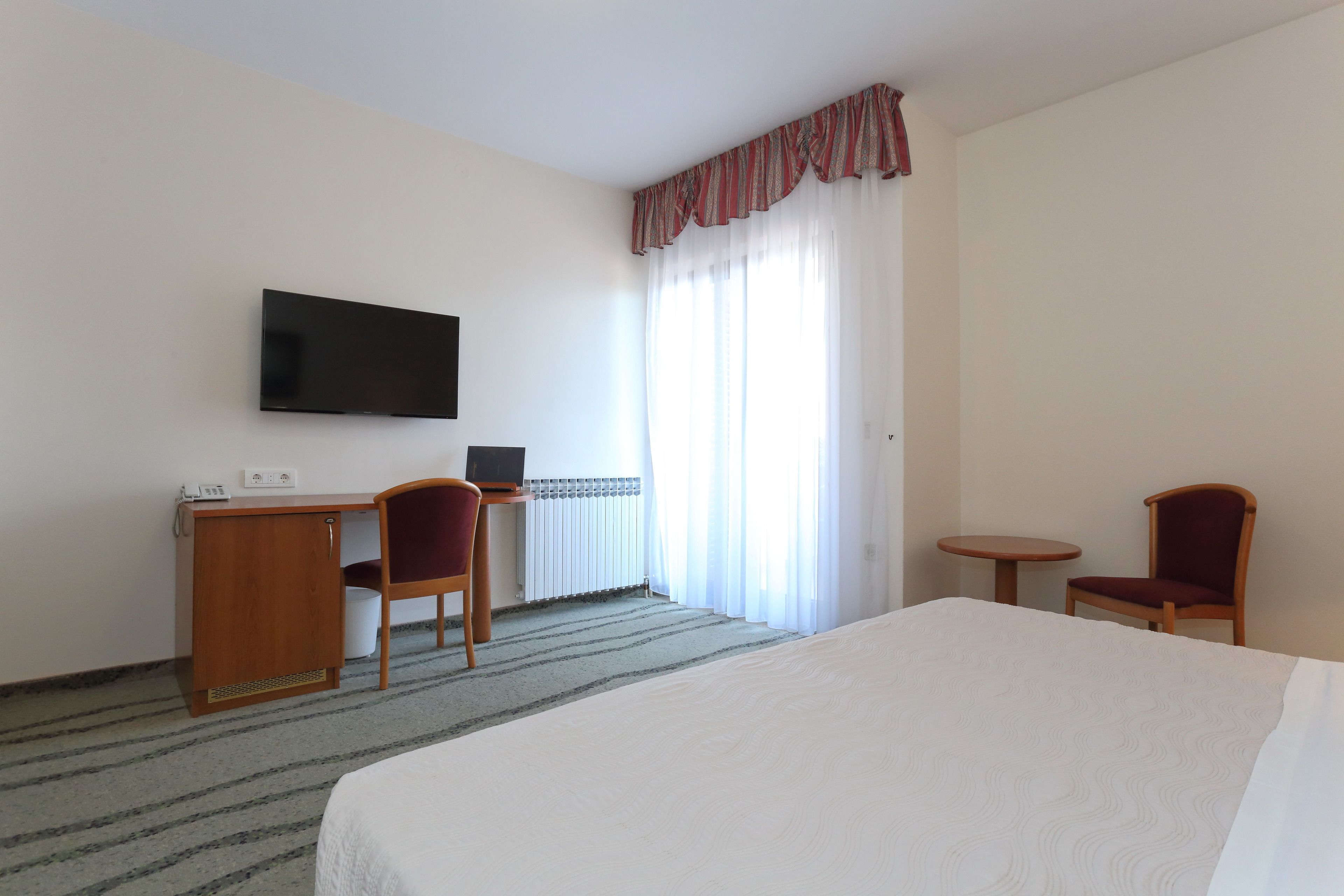 Quarto de hotel com cama, secretária com cadeira, televisão e janela com cortinas.