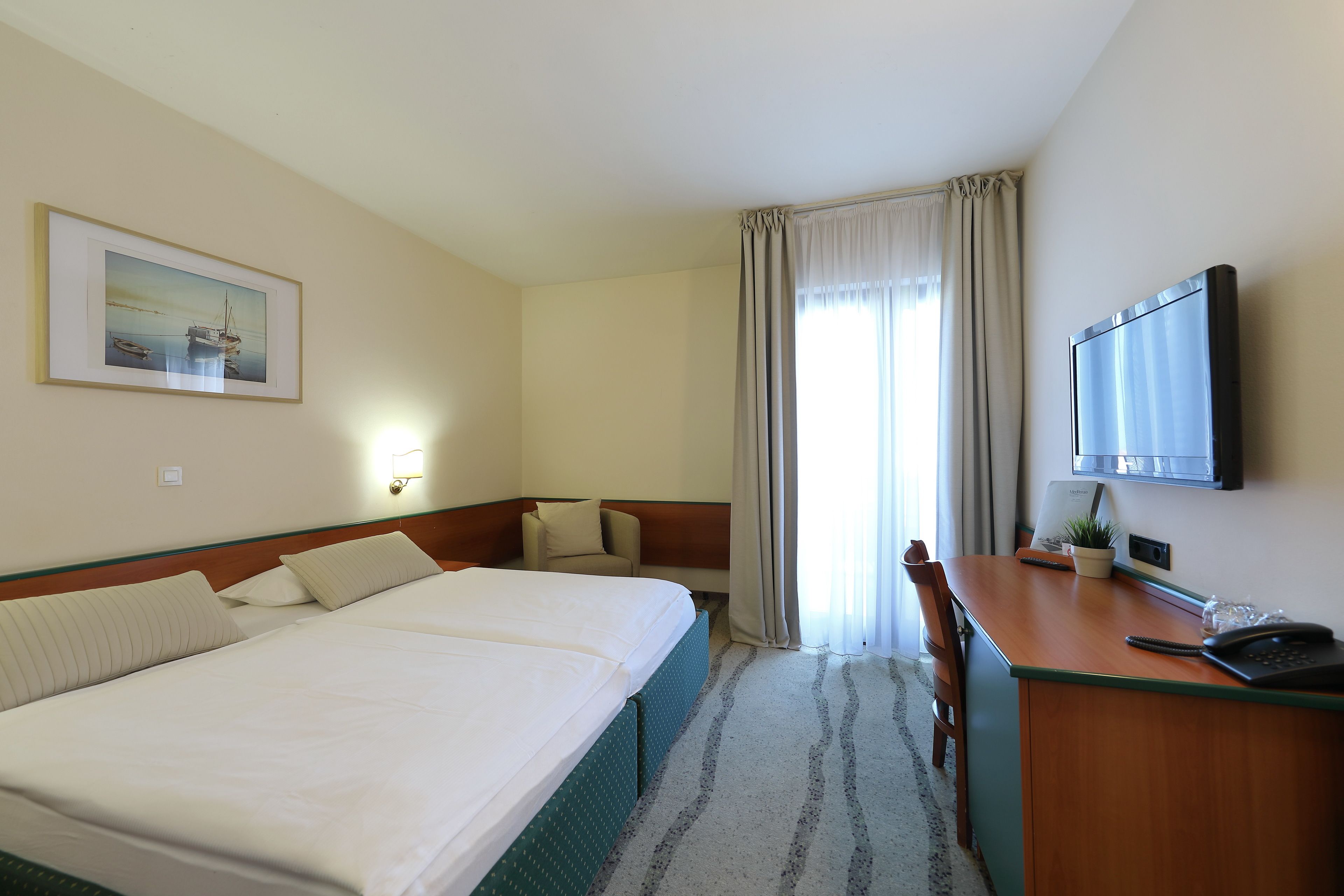Quarto de hotel com uma cama grande, uma escrivaninha com computador, televisão e uma janela com cortinas.