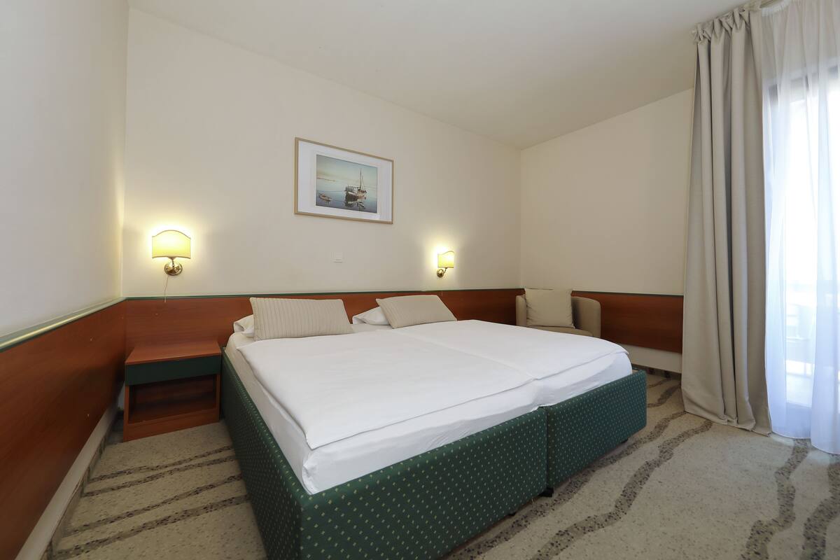 Quarto de hotel com uma cama, duas luminárias fixadas na parede, um quadro e uma janela com cortinas.