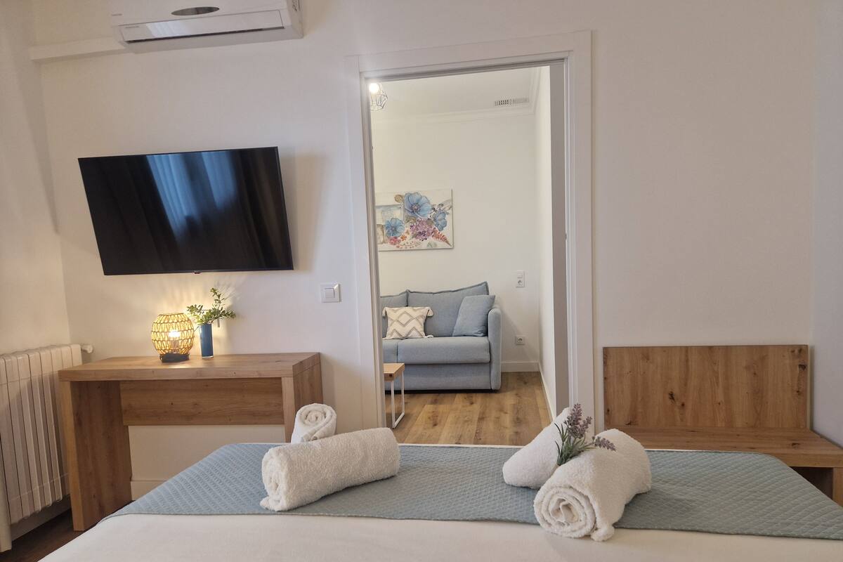 Quarto de hotel com uma cama, uma TV, uma mesa de cabeceira com abajur e vista para outro quarto com sofá.