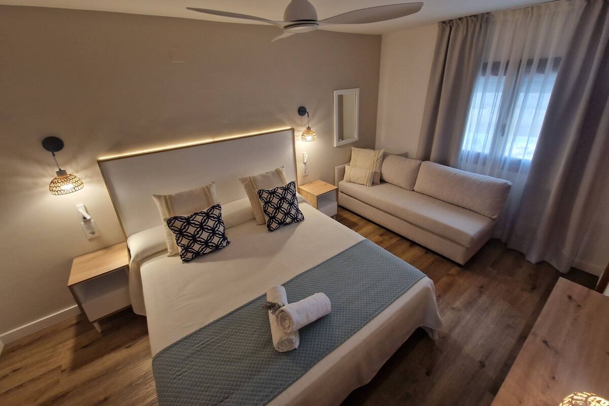 Quarto de hotel moderno com uma cama grande, um sofá, um ventilador de teto e uma janela com cortinas.