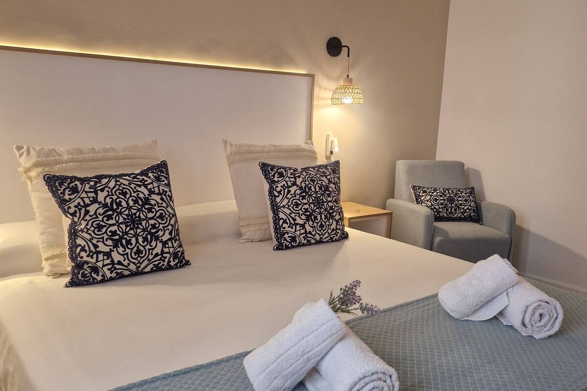 Quarto de hotel com uma cama grande, duas mesas de cabeceira, uma cadeira e uma luminária fixada na parede.