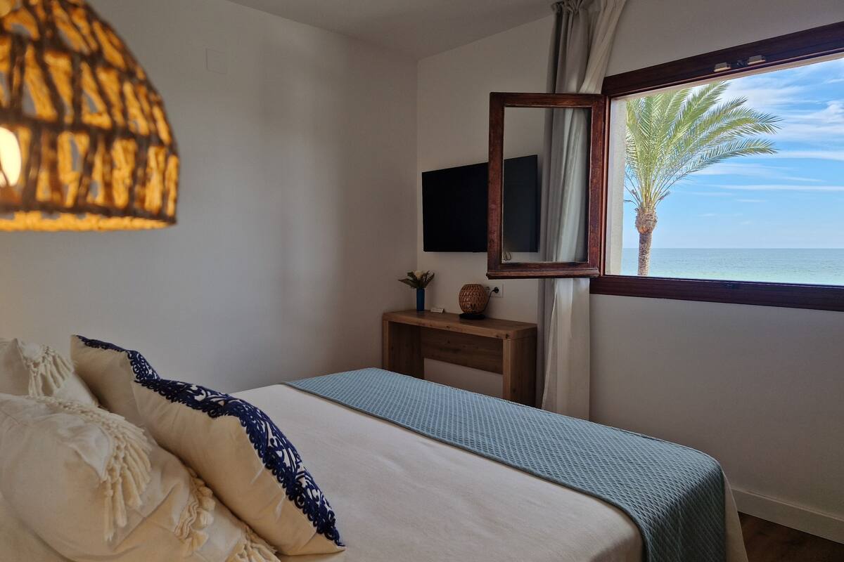 Quarto com cama, mesa de cabeceira, espelho e vista para o mar.