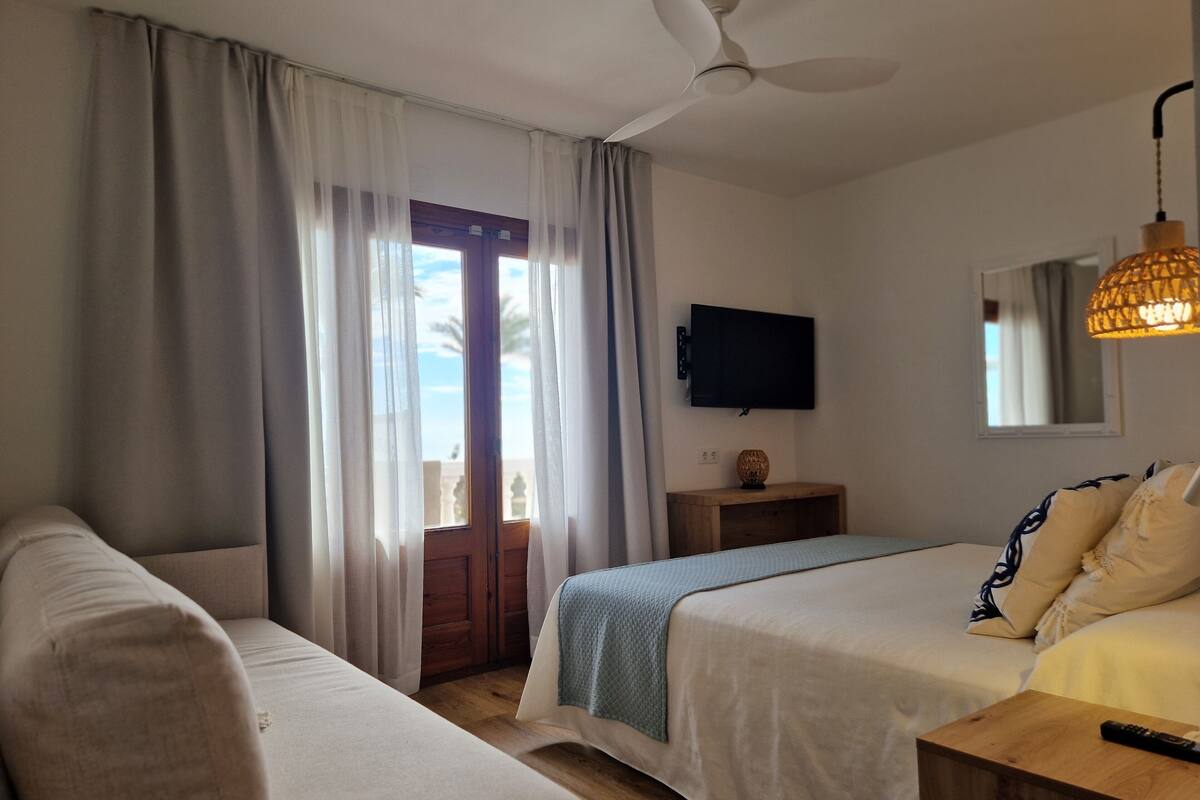 Quarto de hotel com cama, sofá, televisão, ventoinha no teto e janela com cortinas.