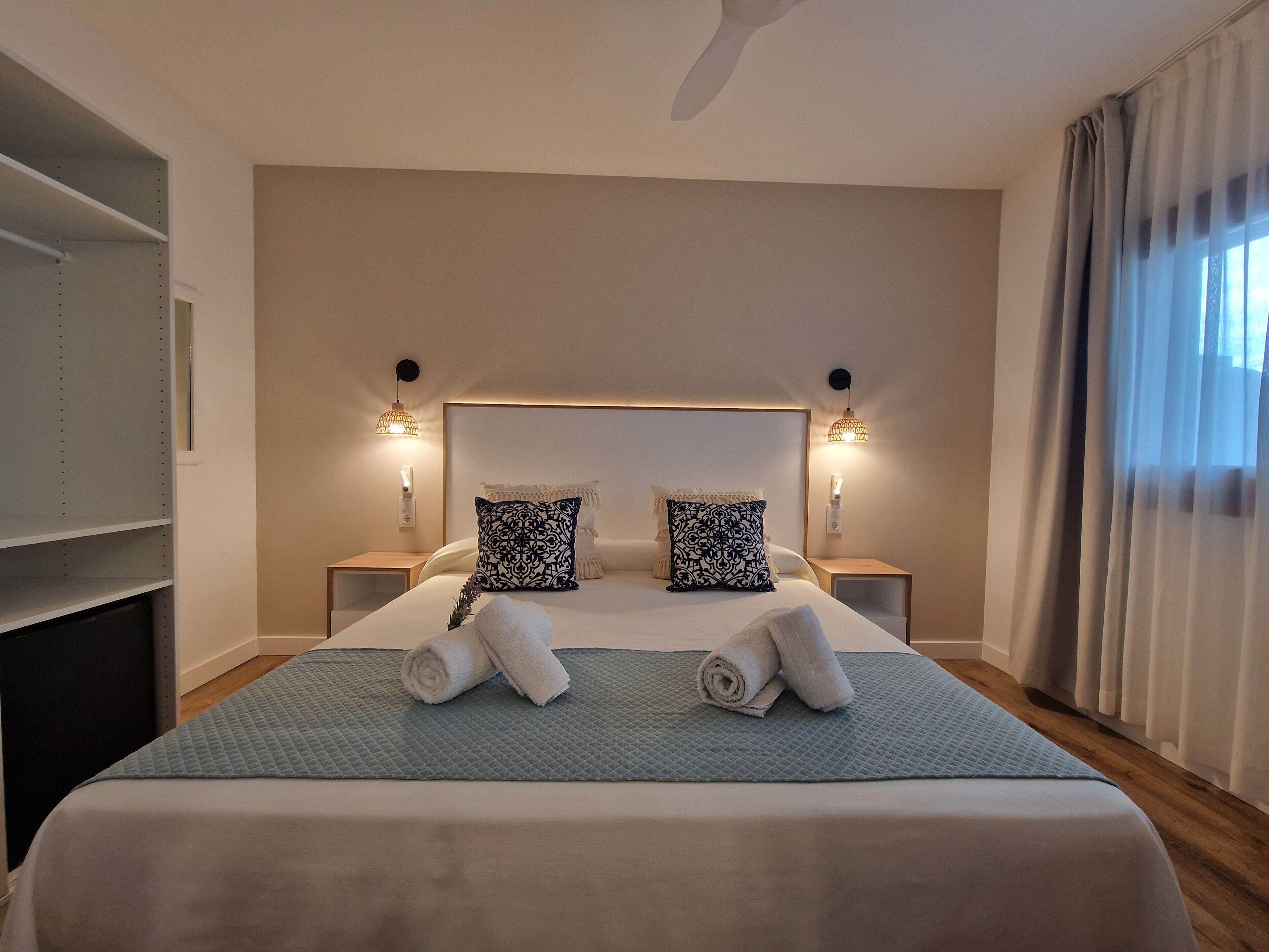 Uma cama bem arrumada com duas almofadas, uma mesa de cabeceira com abajur e uma janela grande com cortinas.