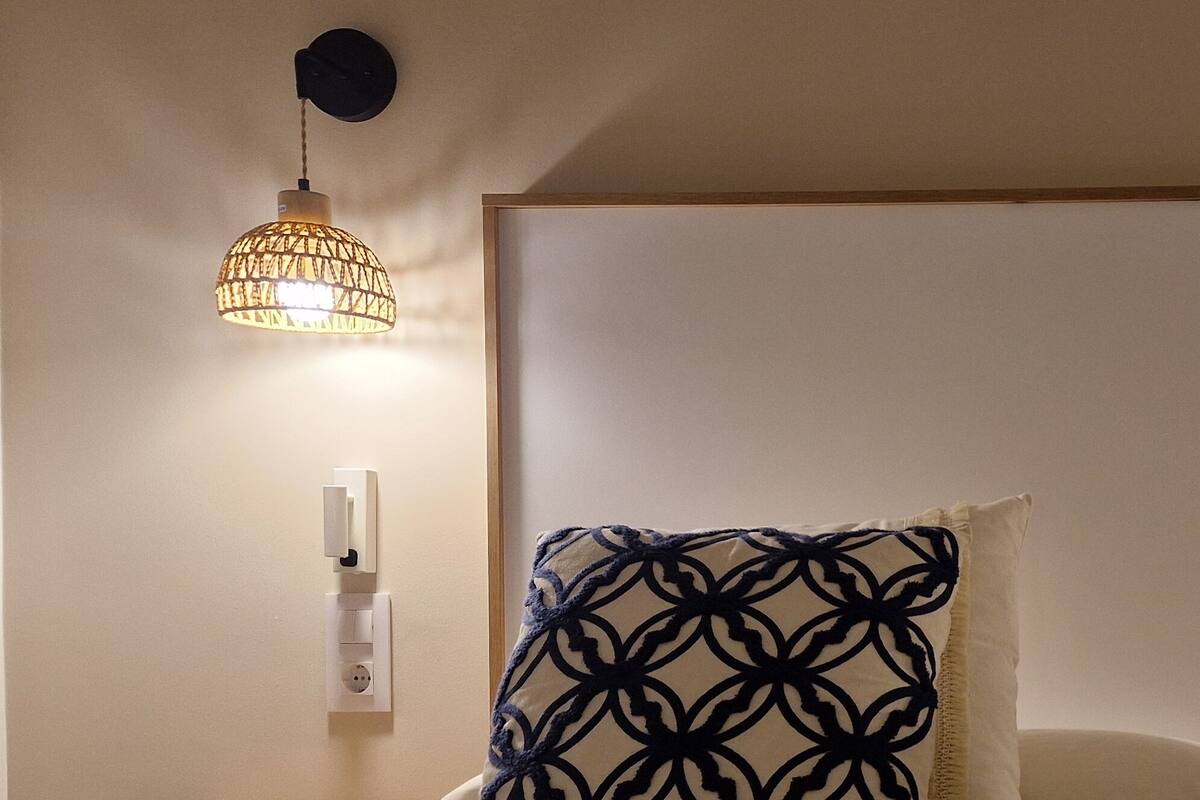 Cama bem arrumada com travesseiro estampado, luminária de cabeceira e tomada na parede.