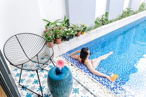 Mulher relaxando perto de uma piscina, com plantas em vasos e uma cadeira.