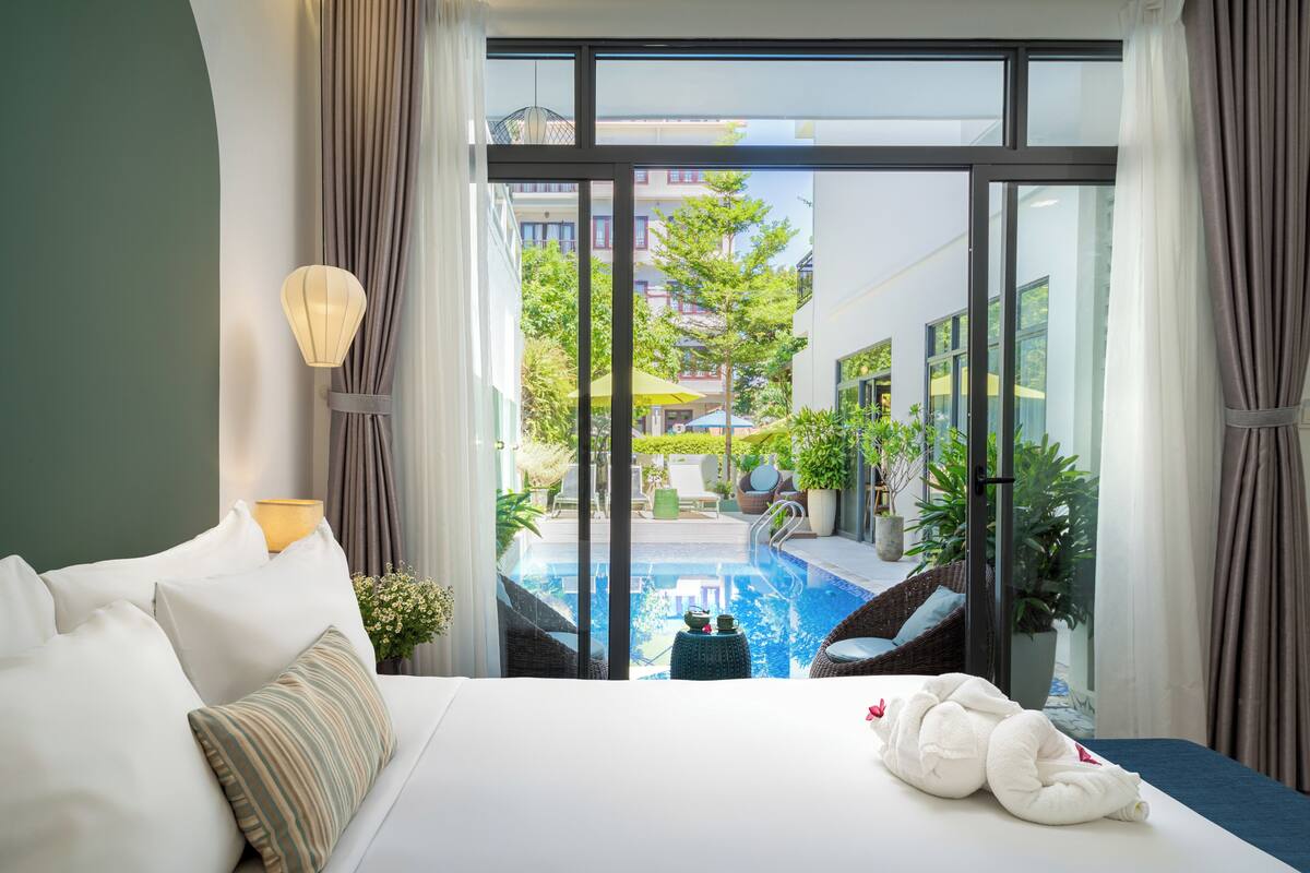 Quarto de hotel com cama, vista para uma piscina e varanda com mobiliário externo.