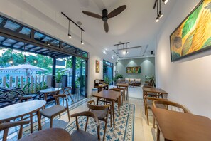 Um café moderno com mesas e cadeiras de madeira, um ventilador de teto e grandes janelas com vista para o exterior.