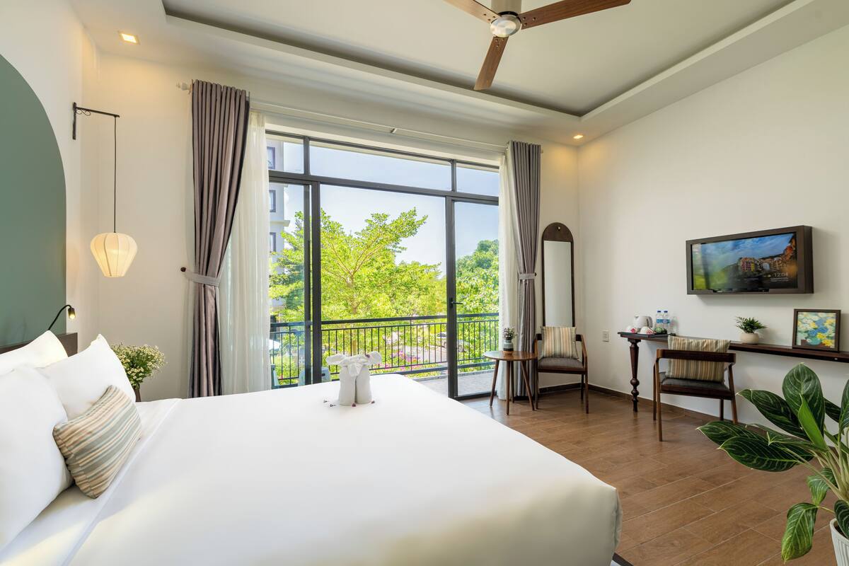 Quarto de hotel com uma cama grande, um ventilador de teto, varanda com vista para vegetação, uma pequena mesa com uma cadeira e uma televisão na parede.