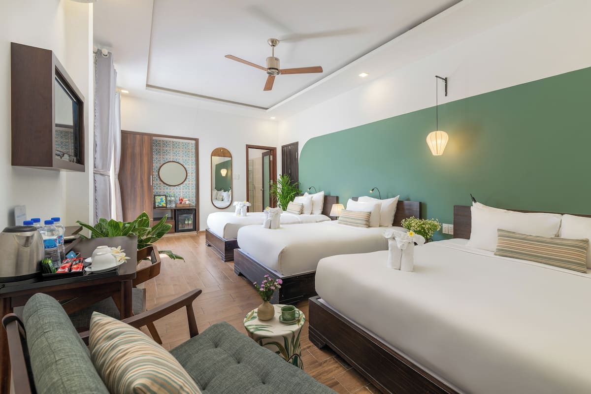 Quarto de hotel com duas camas, escrivaninha com produtos de higiene pessoal, ventoinha no teto e uma parede verde decorativa.