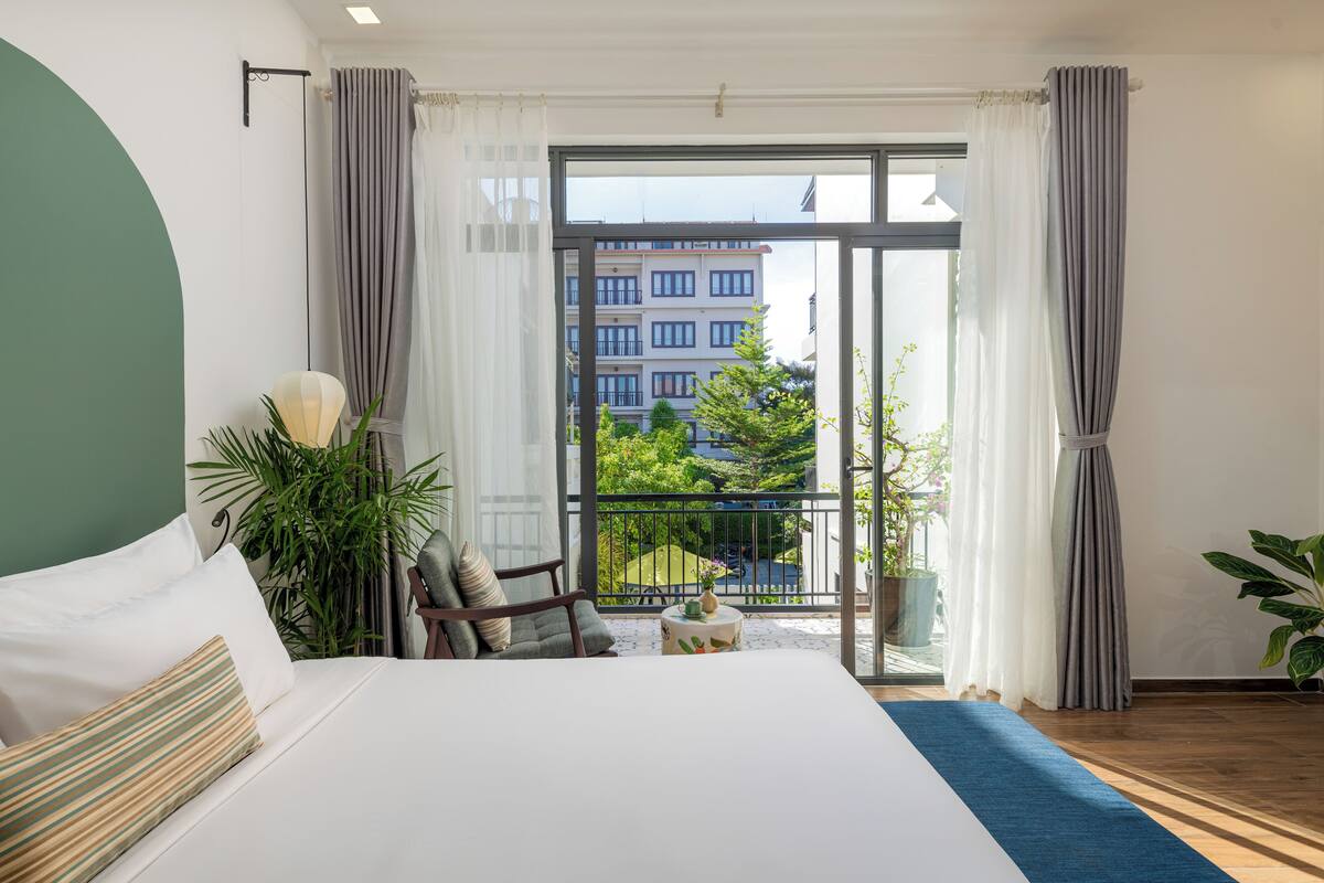 Quarto de hotel com uma cama grande, varanda com vista para edifícios e áreas verdes, uma cadeira e uma pequena mesa com um vaso.