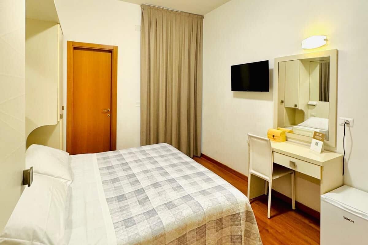 Quarto de hotel com cama, escrivaninha, espelho, televisão e um pequeno frigorífico.