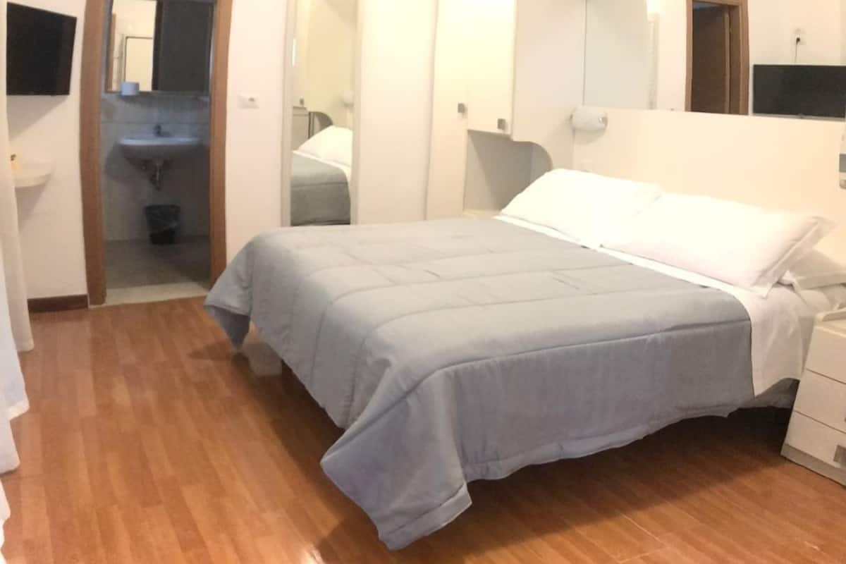 Quarto com cama, piso de madeira, um espelho e porta para um banheiro.