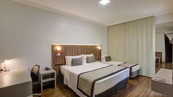 Quarto de hotel com uma cama grande, uma escrivaninha e uma cadeira. Há uma mesa de cabeceira com uma lâmpada e um telefone. O quarto possui piso de madeira e uma janela grande com cortinas.