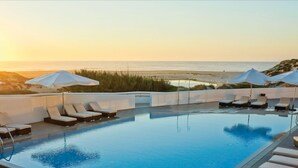 Piscina infinita no terraço com espreguiçadeiras e guarda-sóis, com vista para uma praia e oceano ao pôr do sol.