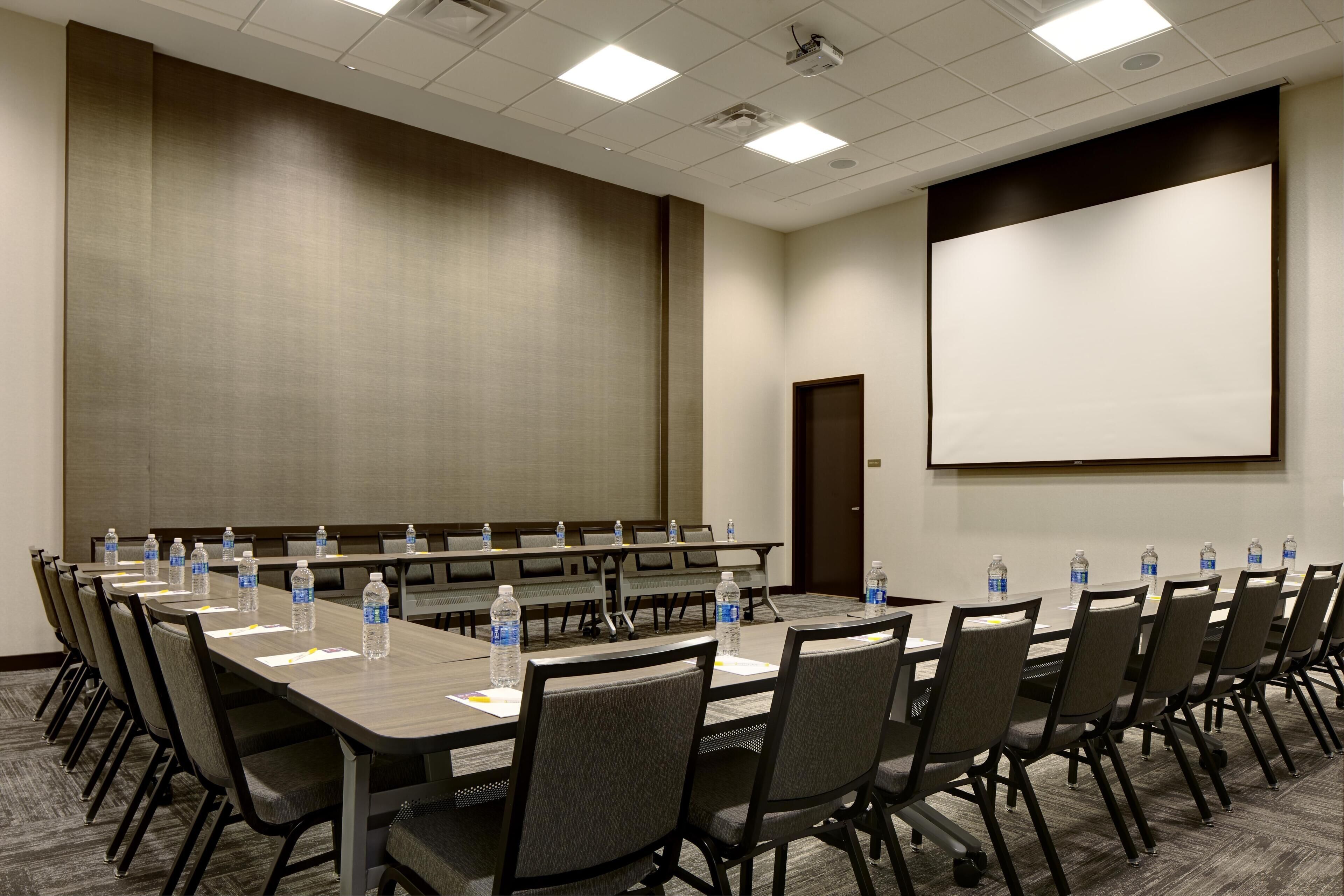 Sala de conferências com uma longa mesa, cadeiras, garrafas de água, tela de projetor e uma porta.