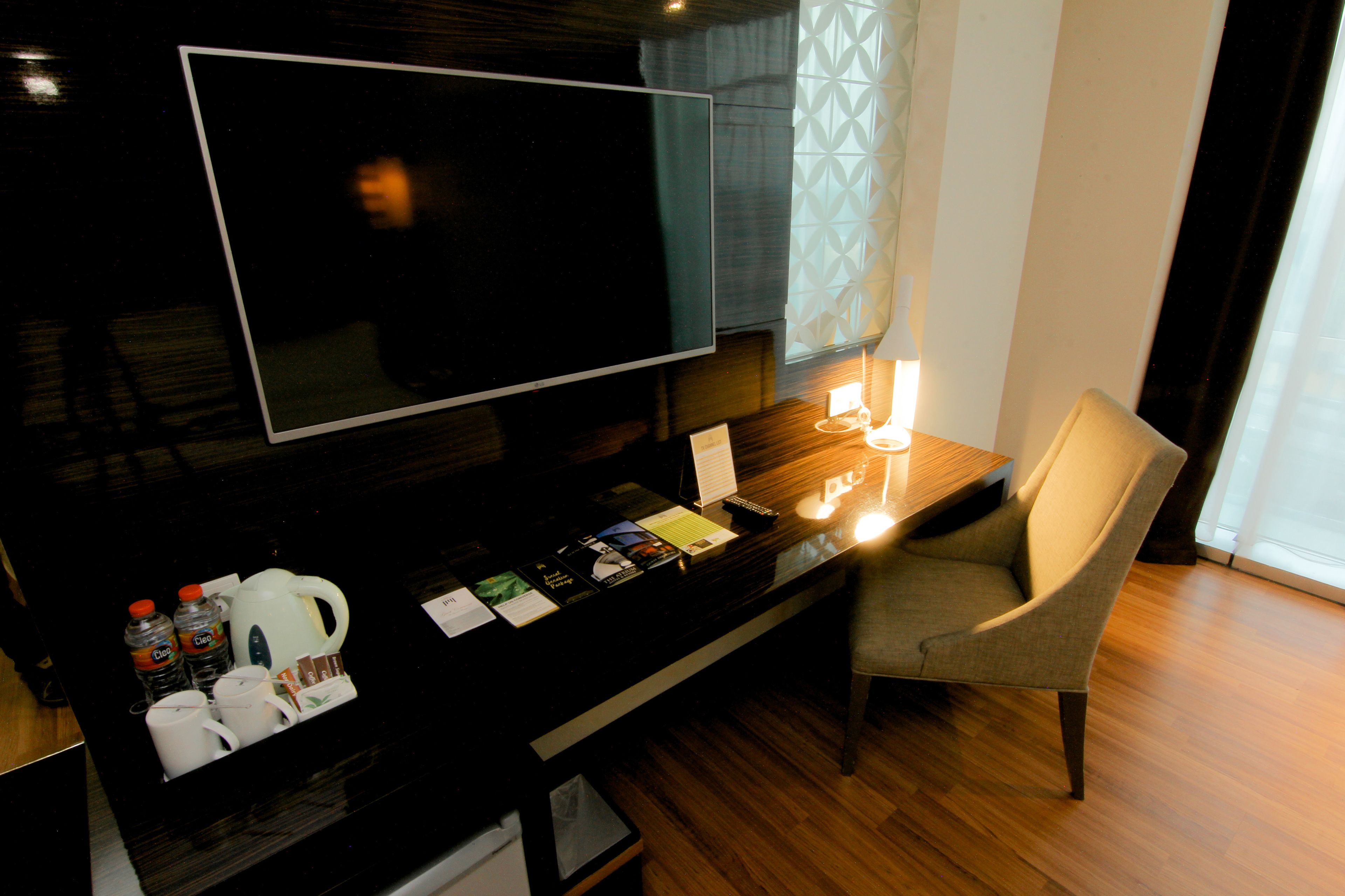 Quarto de hotel com uma TV de tela plana grande, uma escrivaninha de madeira com uma lâmpada, uma cadeira e uma pequena mesa com chaleira e xícaras.