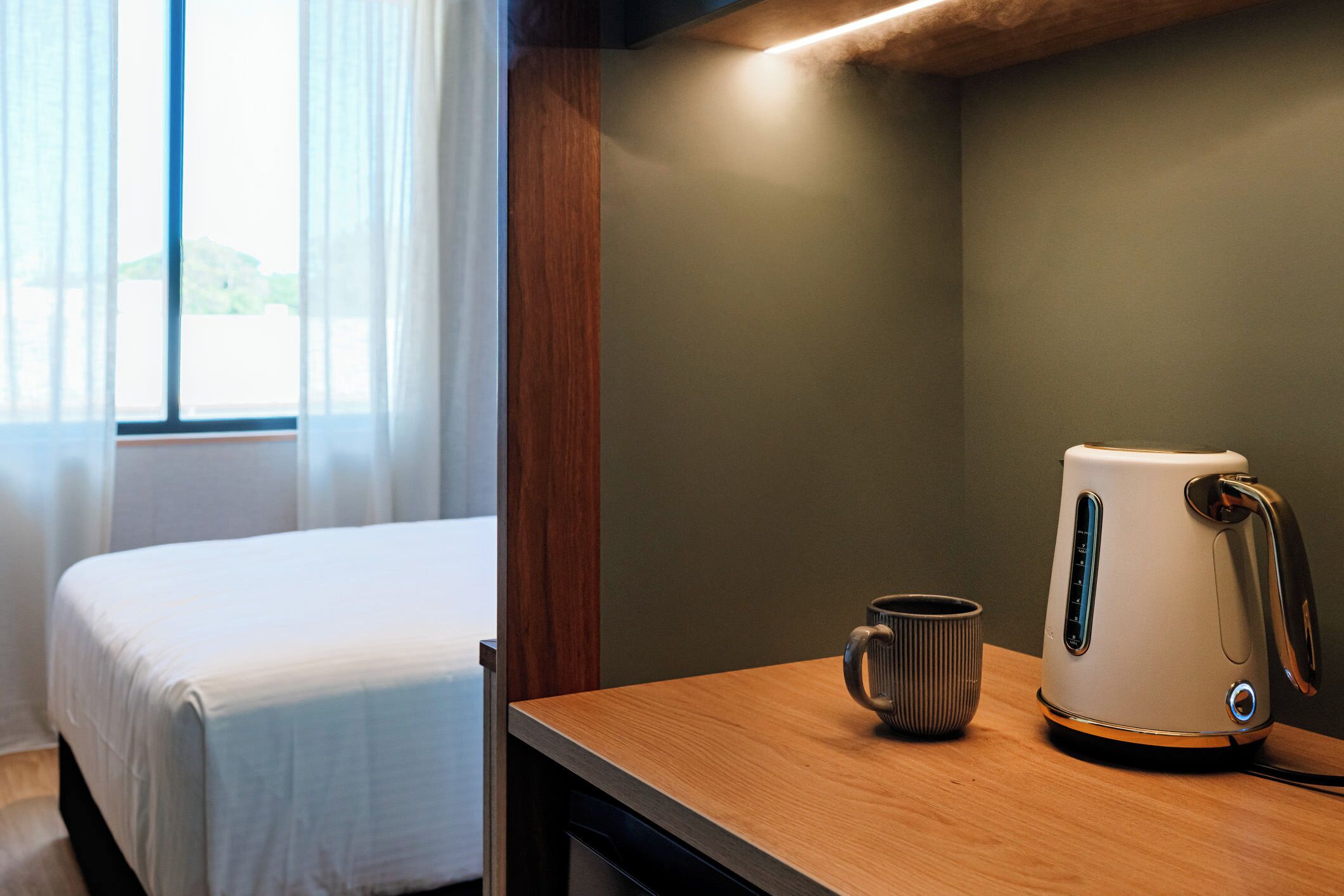 Quarto de hotel com uma cama, uma mesa de cabeceira de madeira, uma chaleira e uma caneca.