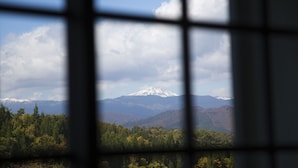 Vista pela janela de uma paisagem montanhosa com pico nevado, colinas arborizadas e céu claro.