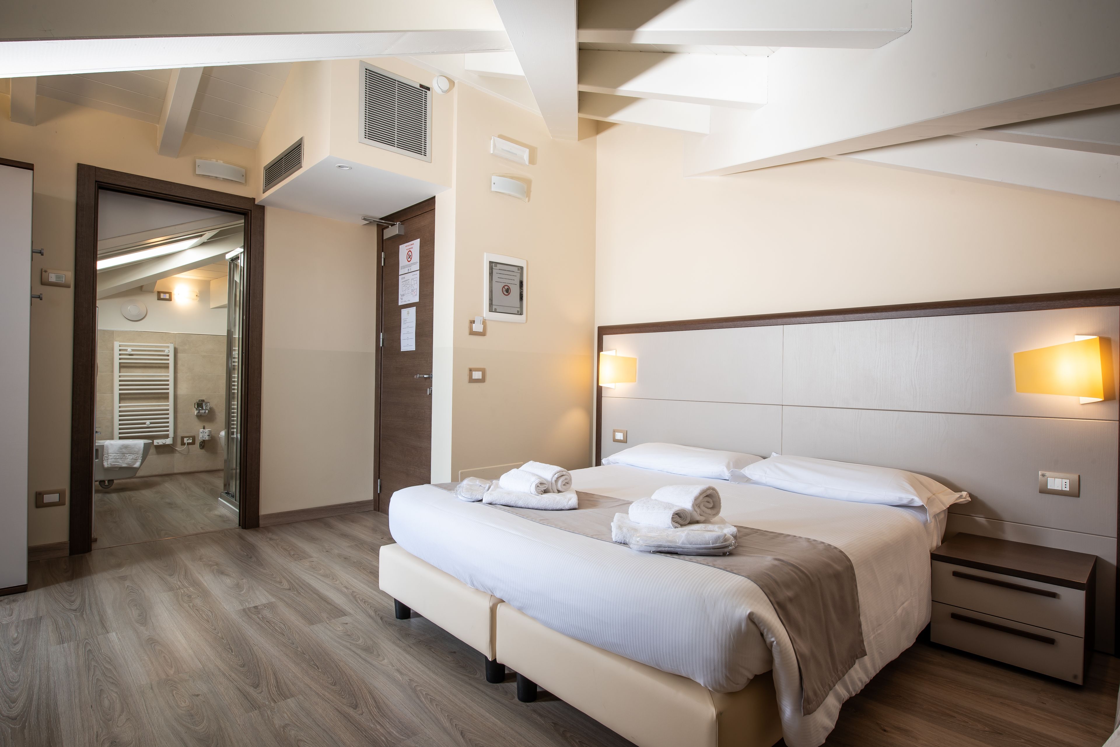 Quarto de hotel com uma cama grande, mesas de cabeceira, um espelho e um banheiro visível através de uma porta aberta.