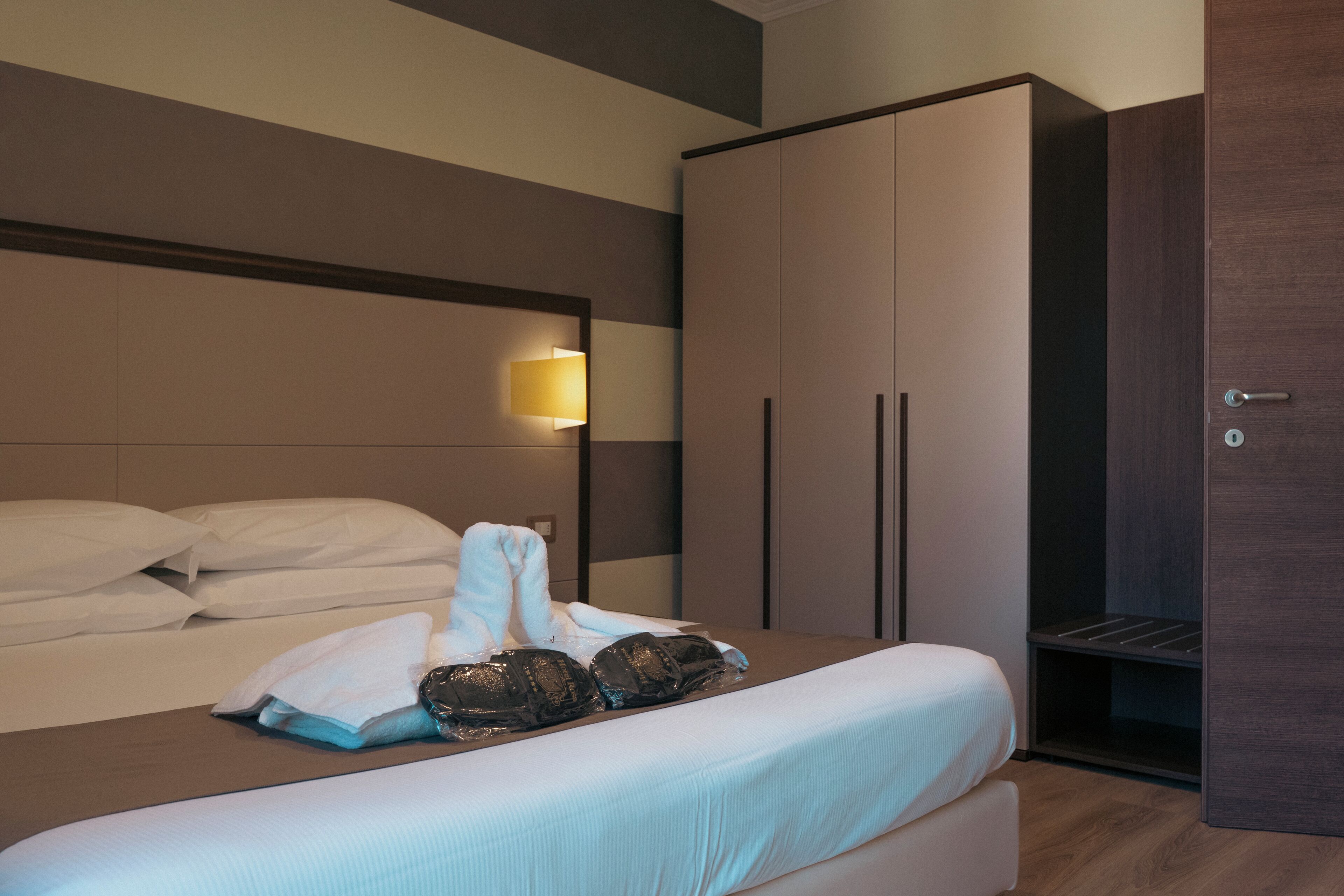 Quarto de hotel com uma cama, um guarda-roupa e uma mesa de cabeceira com abajur.