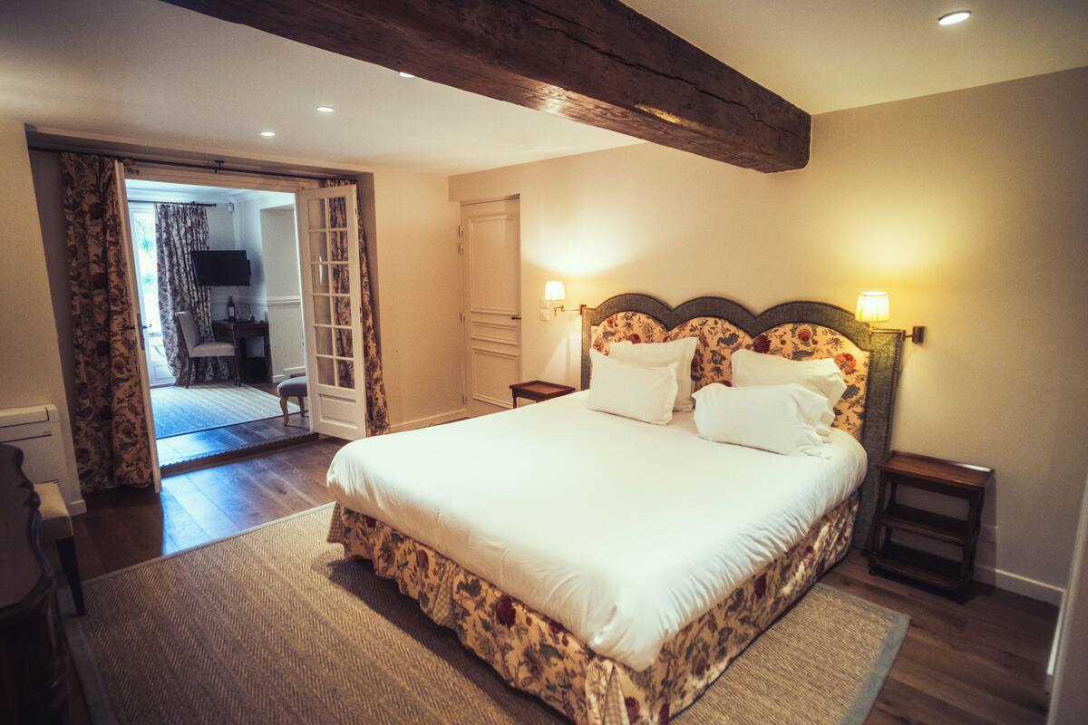 Quarto com uma cama grande, cabeceira de madeira, duas mesas de cabeceira, um criado-mudo, televisão e uma porta para outro cômodo.