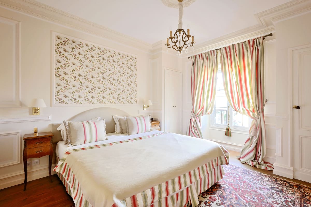 Quarto com cama grande, lustre, cortinas listradas e papel de parede floral.