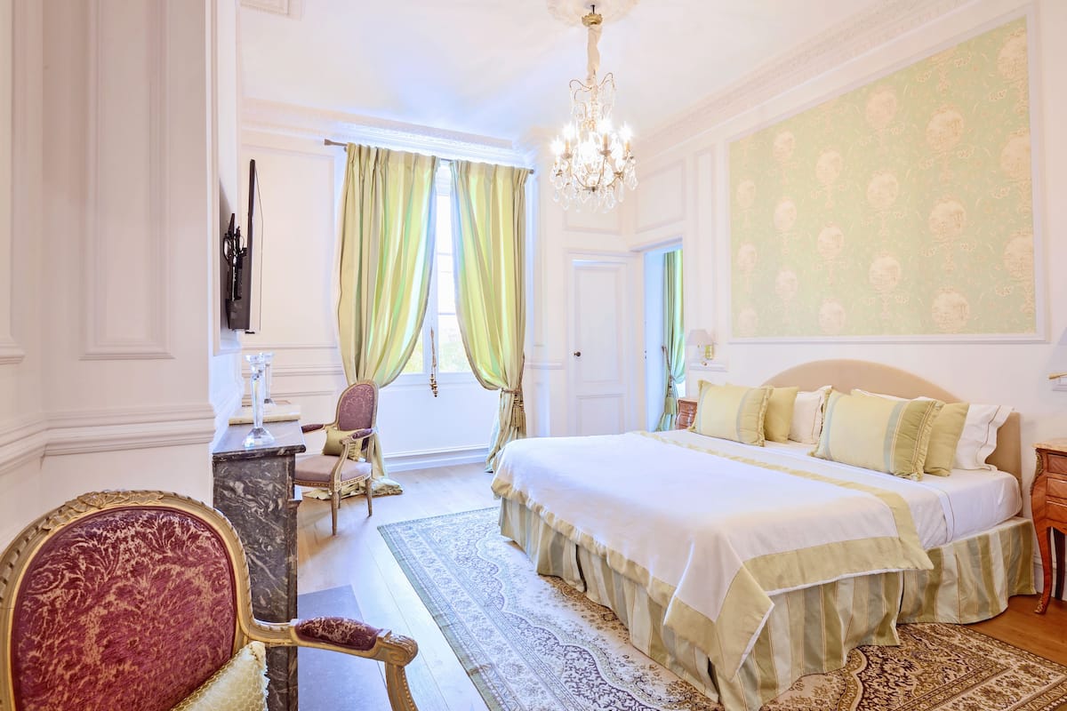 Um quarto espaçoso com uma cama grande, um espaço de estar com uma poltrona e uma mesa, e um lustre.
