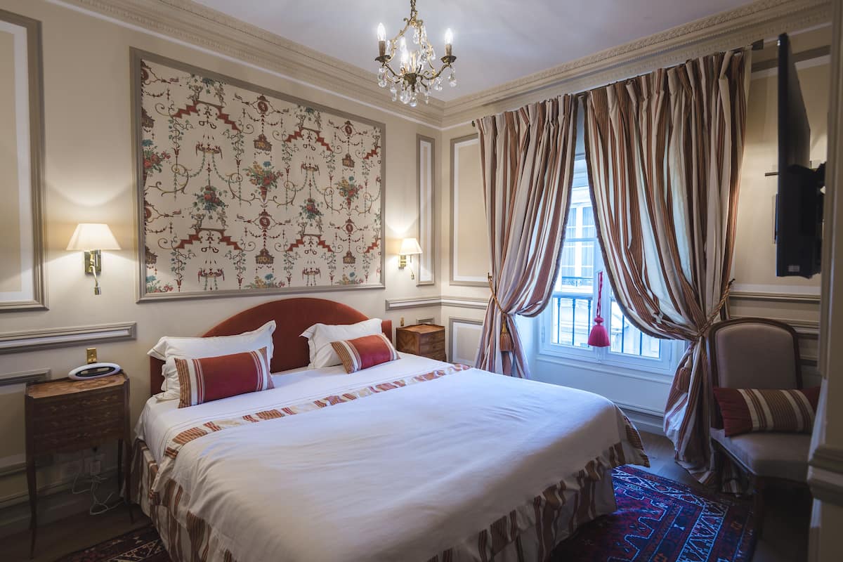 Quarto de hotel com uma cama grande, duas mesas de cabeceira, um candelabro, uma janela ampla com cortinas e uma parede estampada.