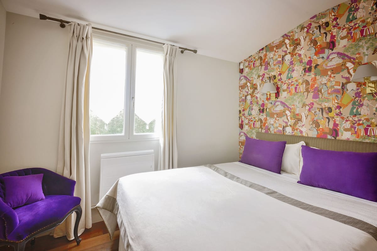 Quarto de hotel com uma cama grande, uma poltrona roxa, uma janela com cortinas e um mural colorido na parede.