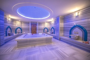 Um spa moderno com uma banheira de hidromassagem central, piso de mármore e arandelas decorativas.