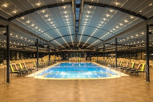 Uma grande piscina interior com teto alto e várias luzes, rodeada por fileiras de espreguiçadeiras.