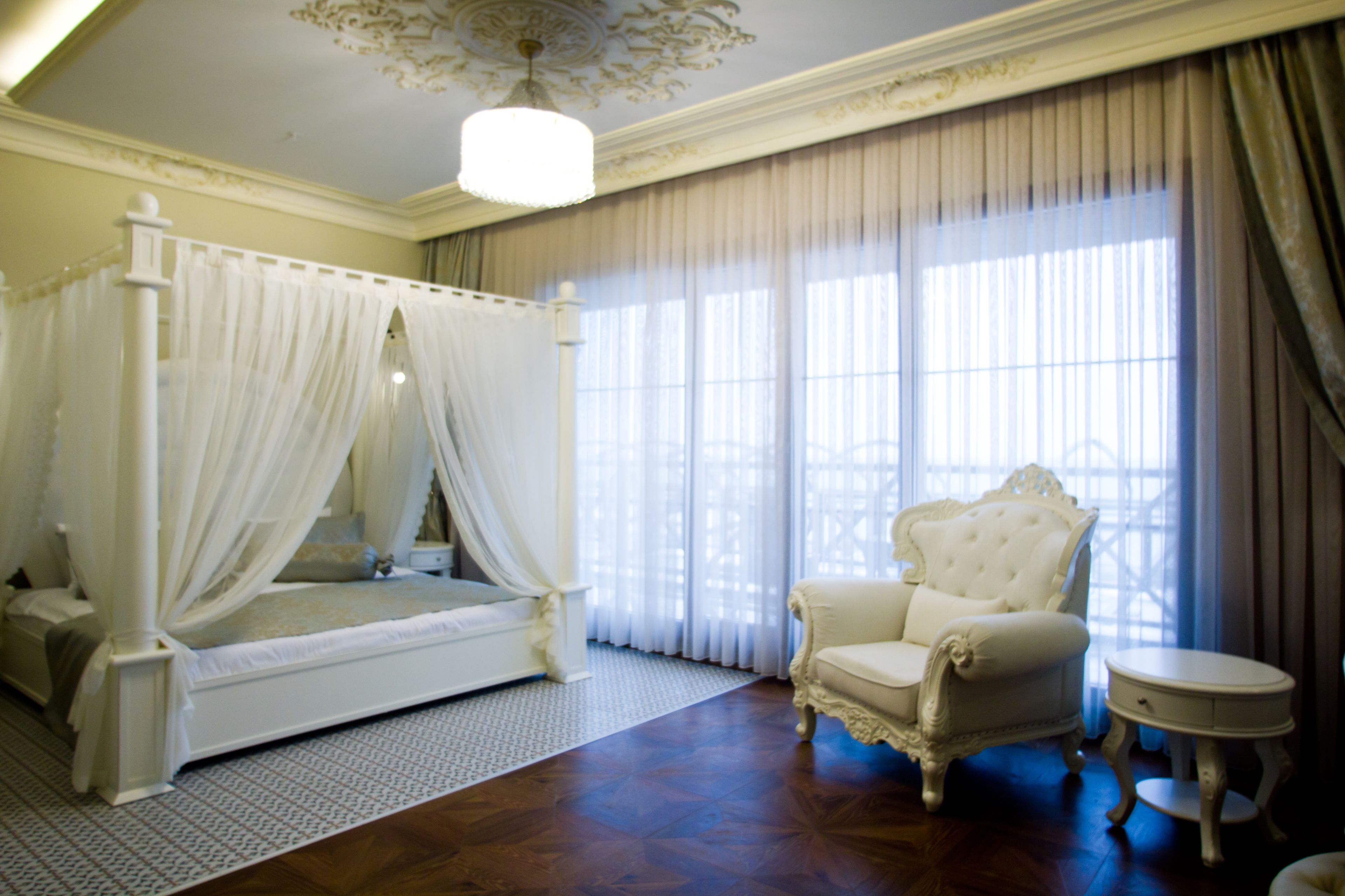 Uma cama de dossel com cortinas brancas, uma poltrona estofada e uma pequena mesa redonda em um quarto com janelas amplas e um candelabro.