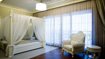 Uma cama de dossel com cortinas brancas, uma poltrona estofada e uma pequena mesa redonda em um quarto com janelas amplas e um candelabro.