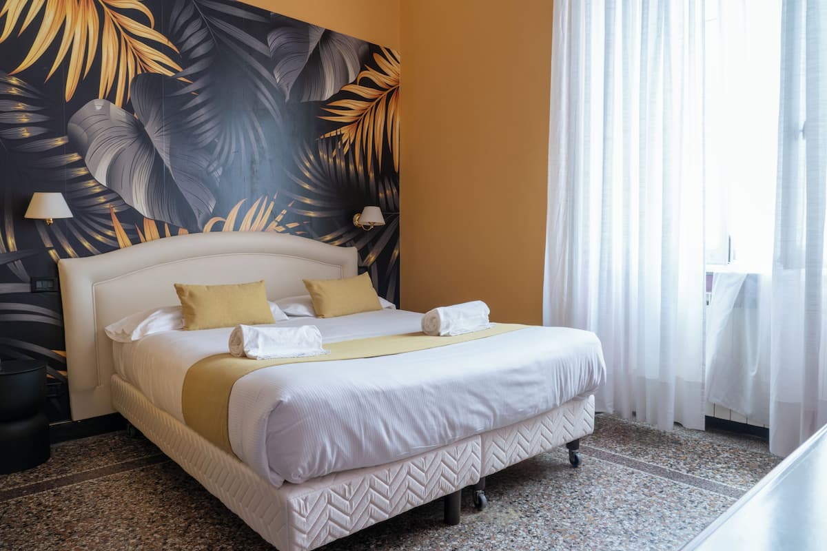 Quarto de hotel com cama, cabeceira, parede com padrão de folhagem tropical, janela com cortinas e abajur em criado-mudo.