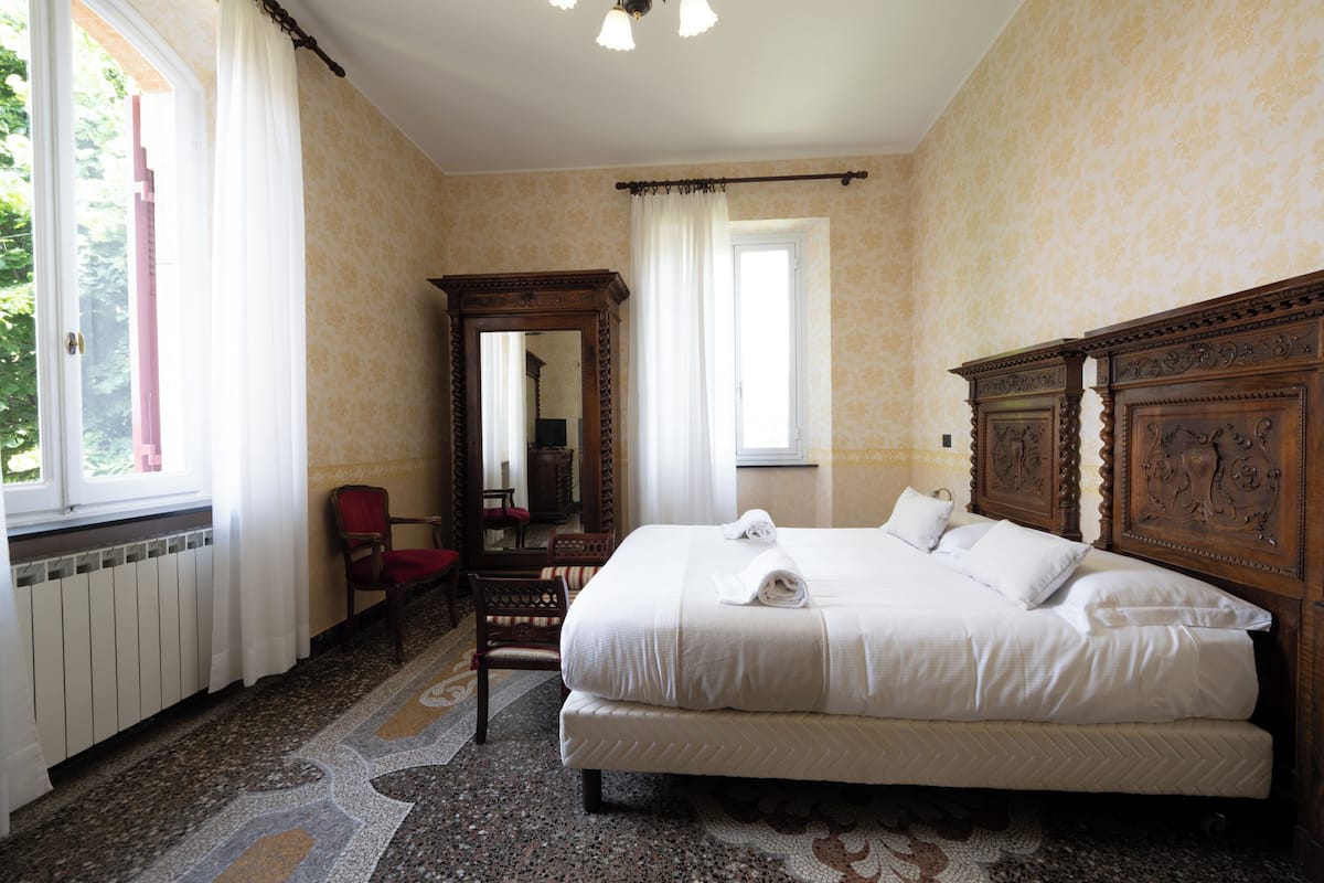 Quarto de hotel com uma cama grande, uma cadeira, um espelho e uma janela com cortinas.