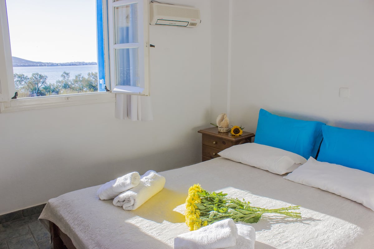 Uma cama bem arrumada com roupa de cama branca, um buquê de flores amarelas e uma janela com vista para um corpo de água e árvores.